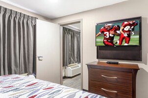 Quarto de hotel com uma cama, televisão fixada na parede, cômoda de madeira e porta que dá para outro quarto.