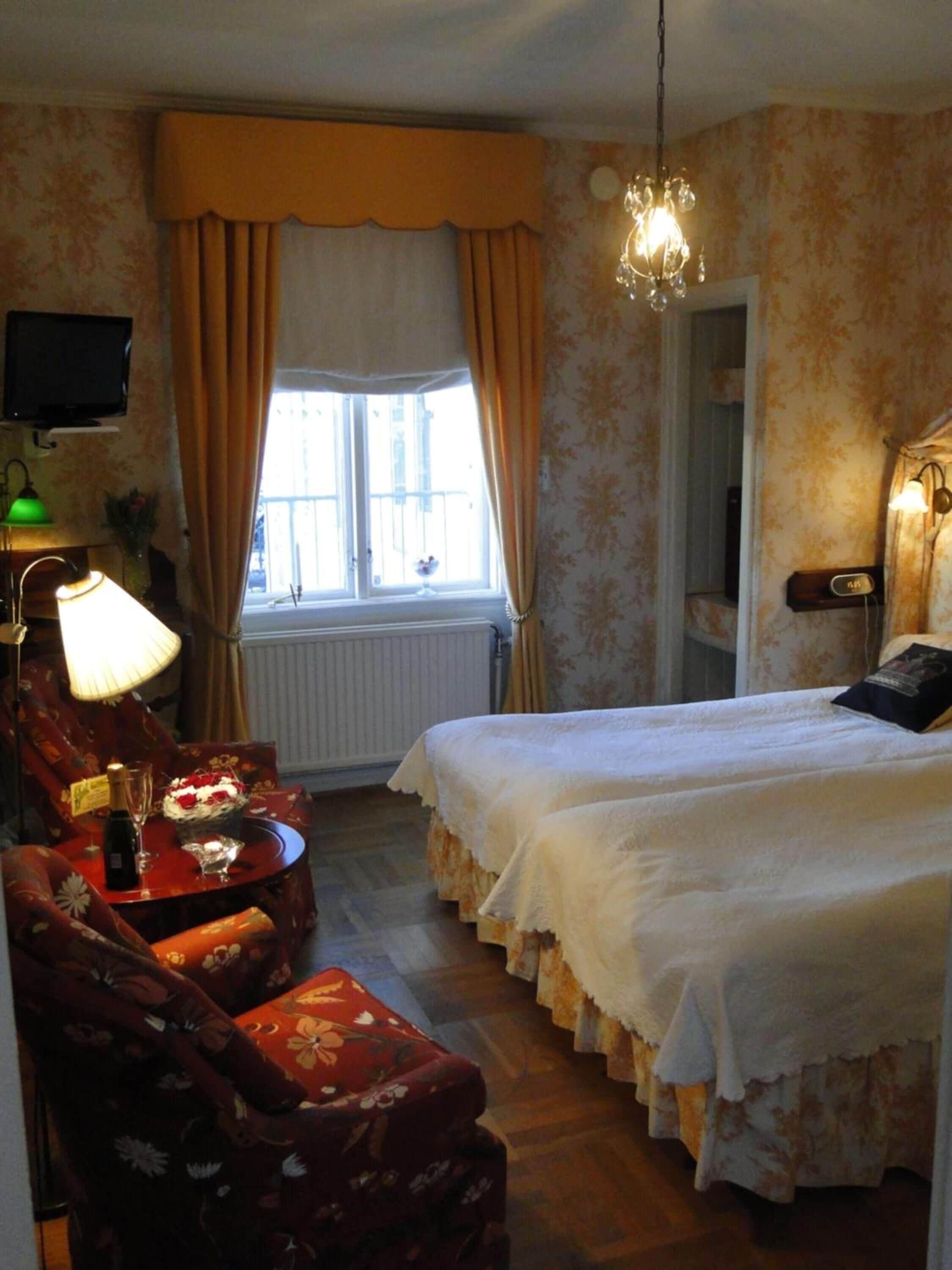 Quarto de hotel com uma cama, uma mesa com luminária, uma cadeira e uma janela com cortinas.