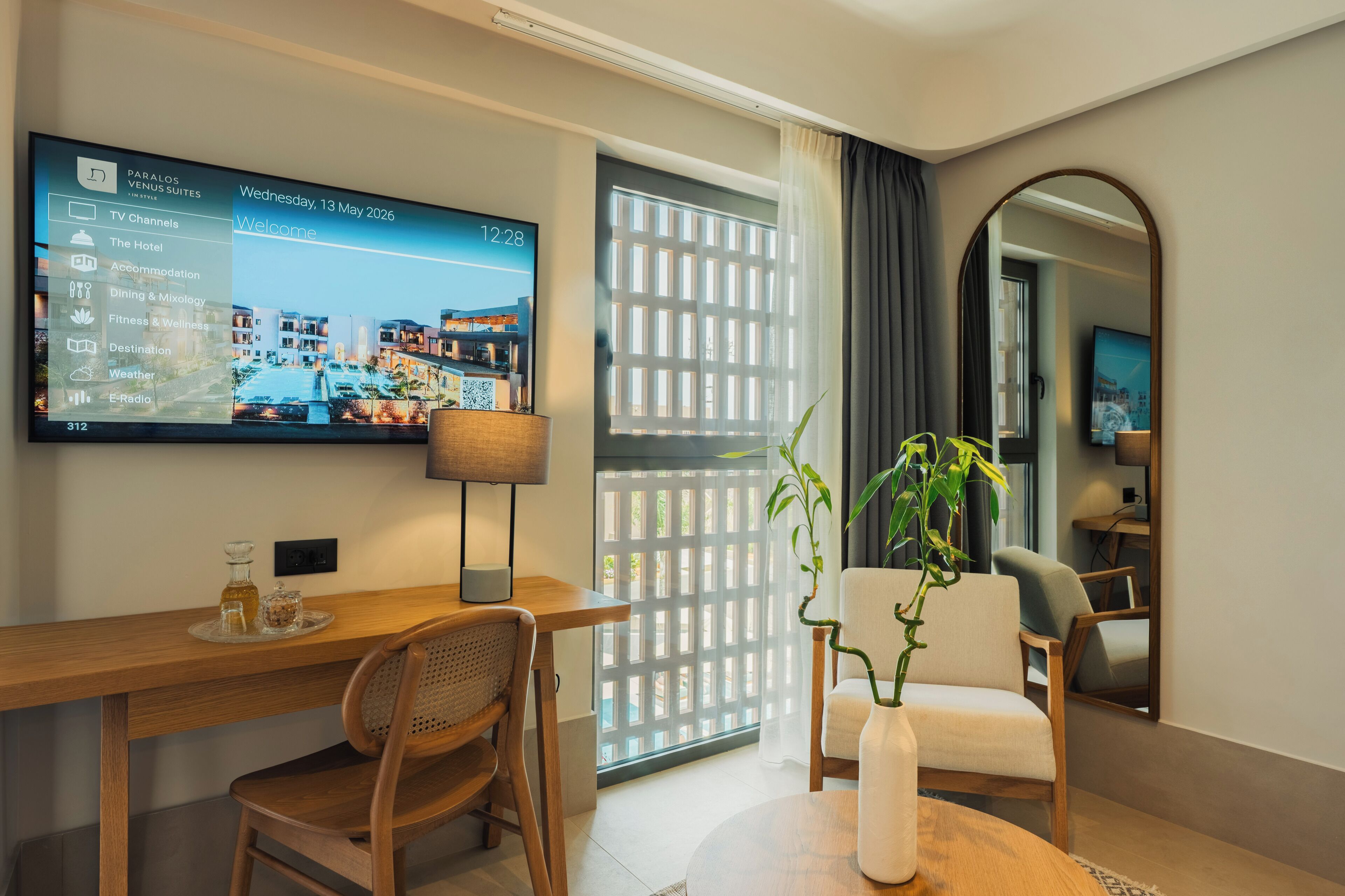 Quarto de hotel com televisão exibindo uma tela de boas-vindas, escrivaninha com cadeira, mesinha com um vaso e uma grande janela com vista para o hotel.