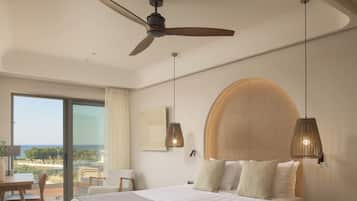 Quarto de hotel moderno com uma cama grande, um ventilador de teto, vista para o oceano e um espaço de refeições com mesa e cadeiras.