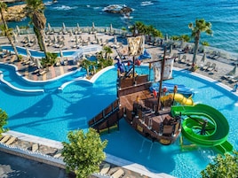 Uma piscina grande ao ar livre com área de recreação temática de navio pirata, rodeada por espreguiçadeiras e palmeiras.