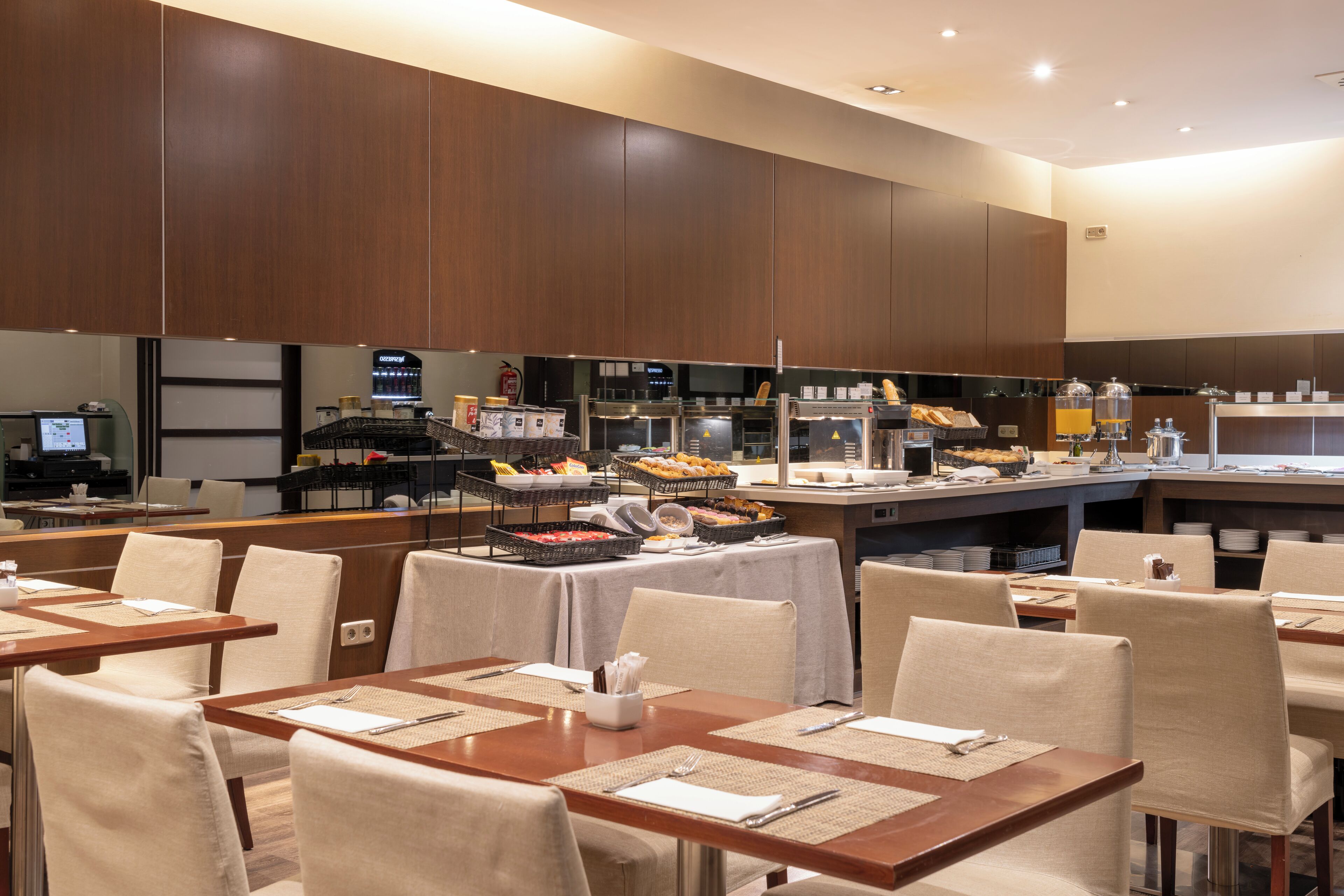 Espaço de refeições de hotel com opções de pequeno-almoço em estilo buffet, disposição de mesas e um canto para café.