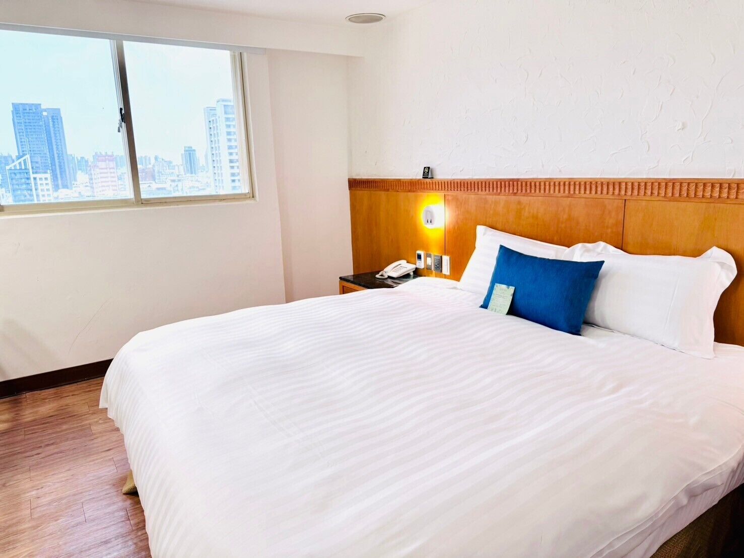 Quarto de hotel com uma cama grande, mesa de cabeceira, telefone e vista da cidade pela janela.
