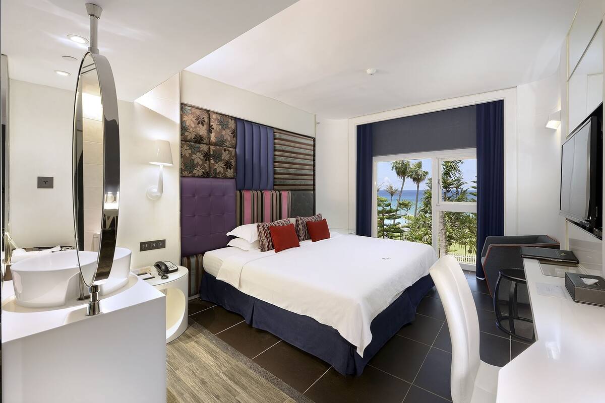 Quarto de hotel moderno com uma cama grande, banheiro com espelho e pia, e vista para o oceano.