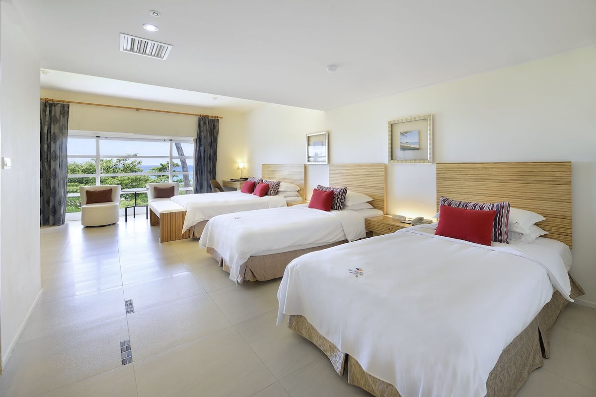 Quarto de hotel com duas camas, área de estar com uma cadeira e mesa, e vista para o exterior.