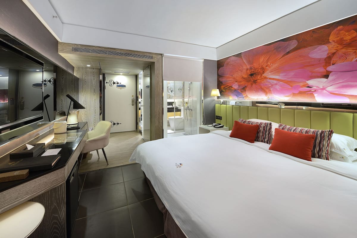Quarto de hotel moderno com uma cama grande, uma escrivaninha com computador, uma cadeira, uma televisão e um banheiro visível através de uma porta aberta.