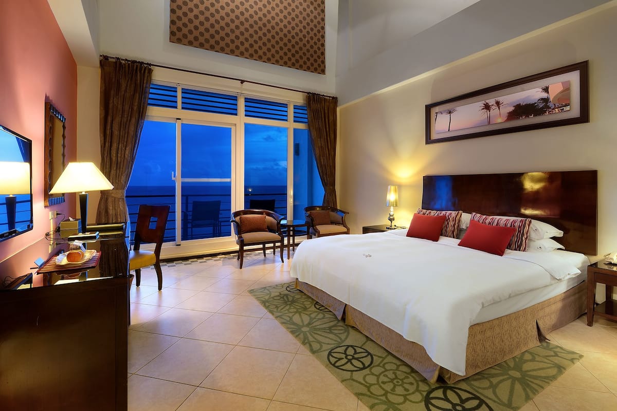 Quarto de hotel com uma cama grande, uma mesa com luminária, uma cadeira, vista para o mar por uma janela ampla e um quadro na parede.