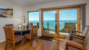 Uma sala de jantar com mesa e cadeiras, um sofá e uma grande porta de vidro deslizante que dá para uma varanda com vista para o mar.