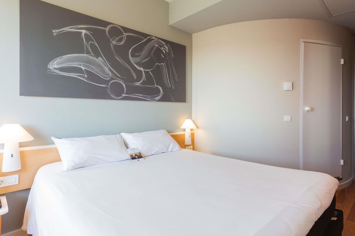 Quarto de hotel com uma cama, duas luminárias de cabeceira, um guarda-roupa e um quadro grande e abstrato na parede.