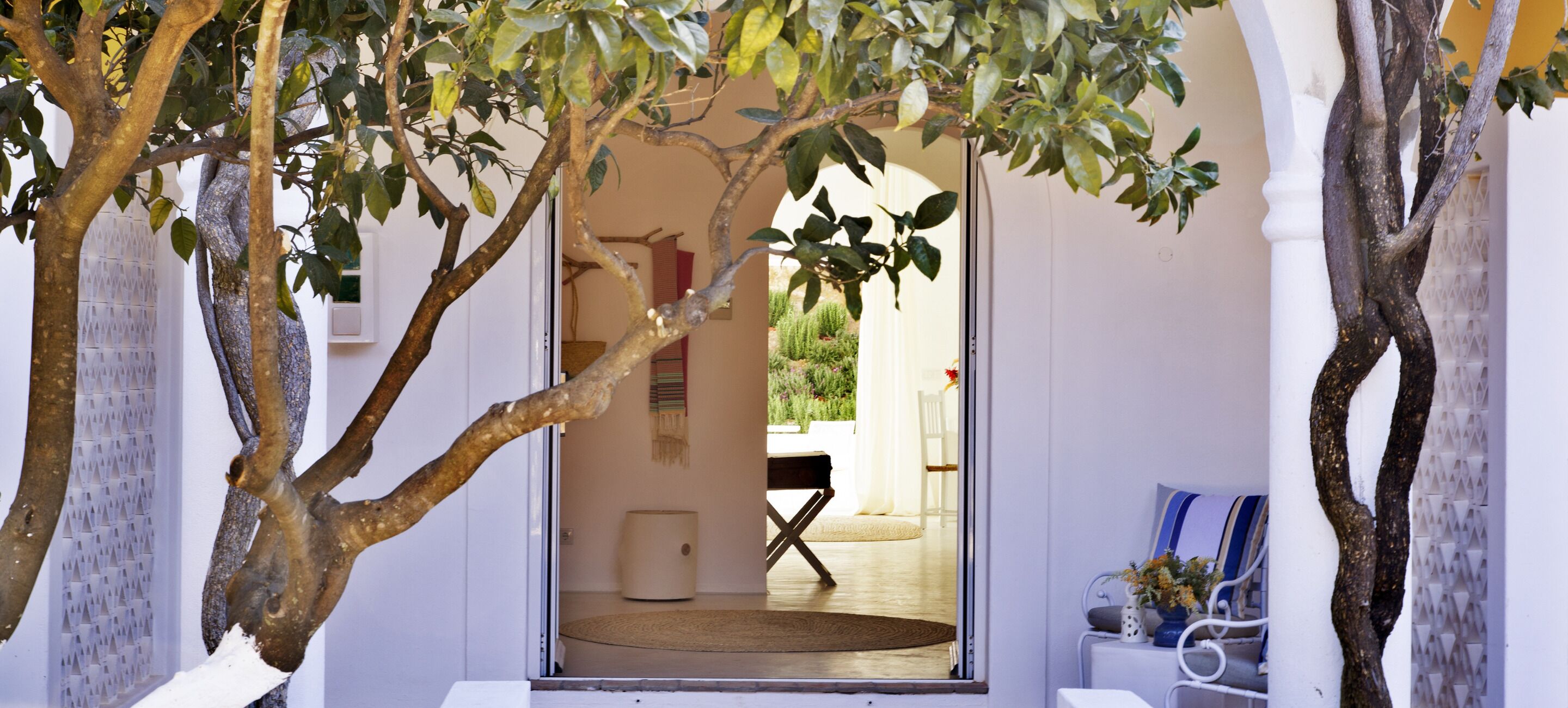 Uma porta arqueada dá acesso a um quarto com um tapete tecido, uma cadeira de madeira e uma pequena mesa com um vaso de flores.