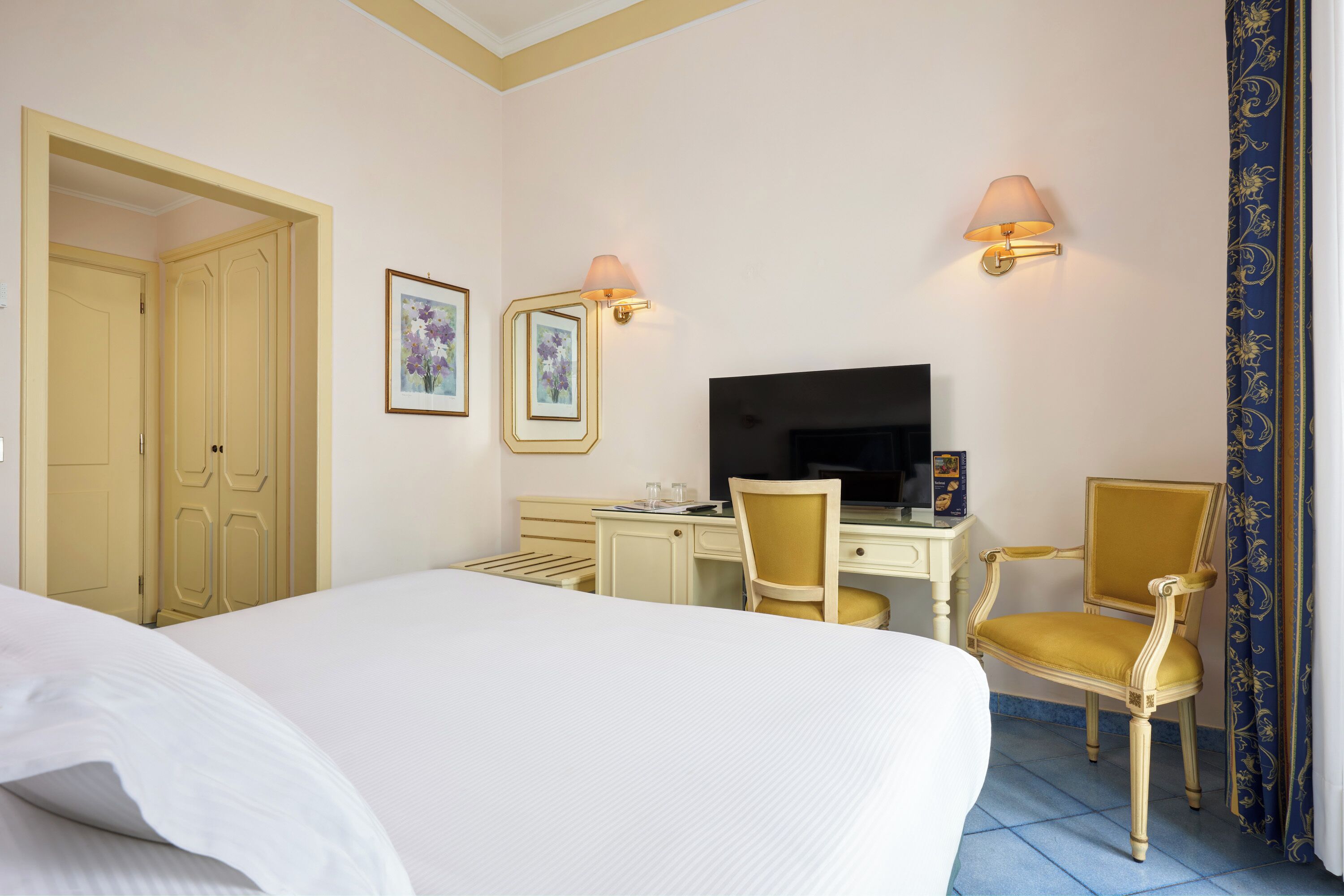 Quarto de hotel com uma cama, escrivaninha, duas cadeiras, televisão e duas luminárias fixadas na parede.