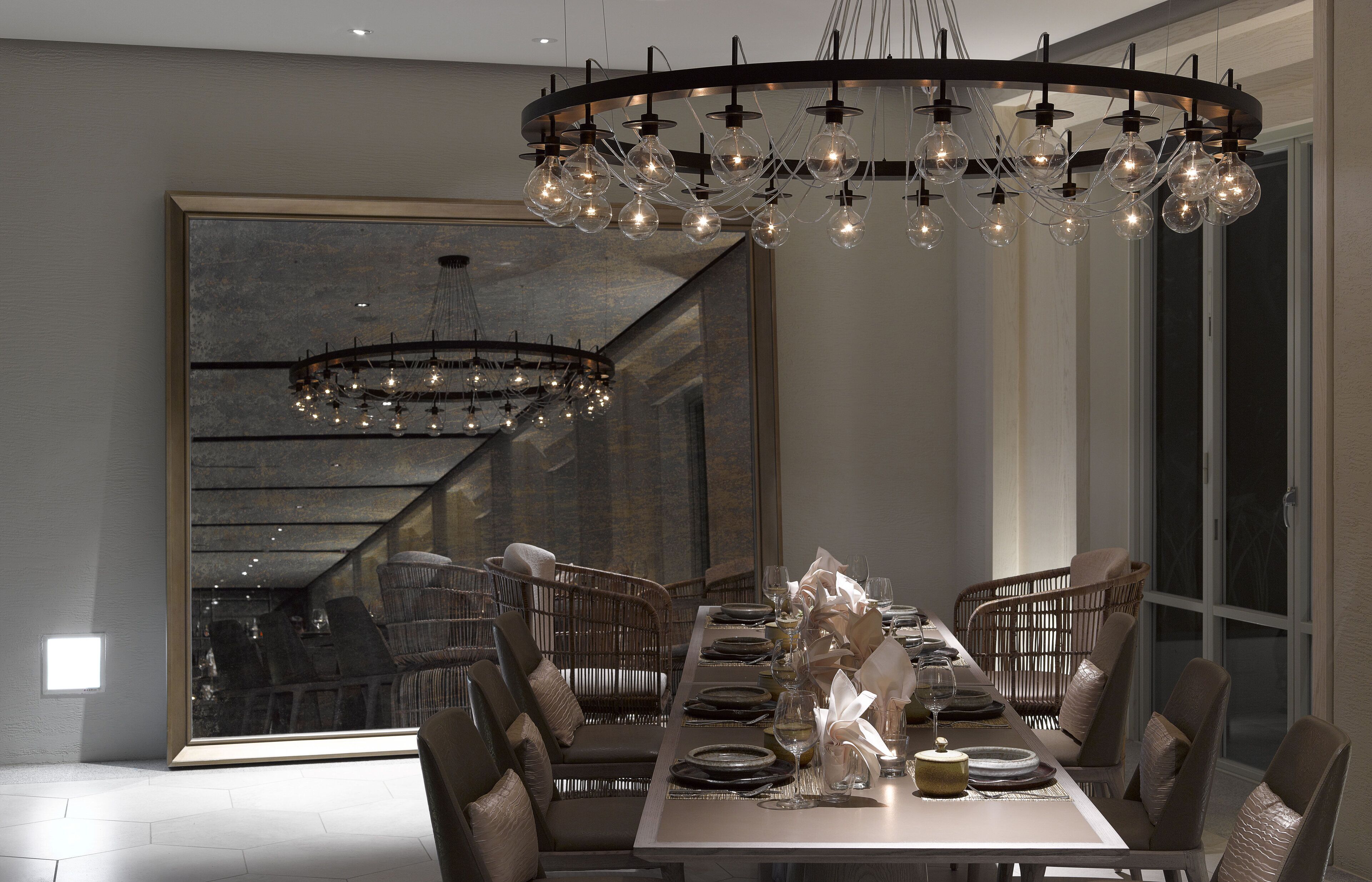 Uma sala de jantar moderna com uma mesa comprida posta para refeição, rodeada por cadeiras de vime, sob um grande candelabro circular.