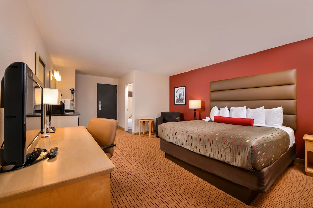 Quarto de hotel com cama, secretária com computador e controles remotos, cadeira, mesinha e abajur.