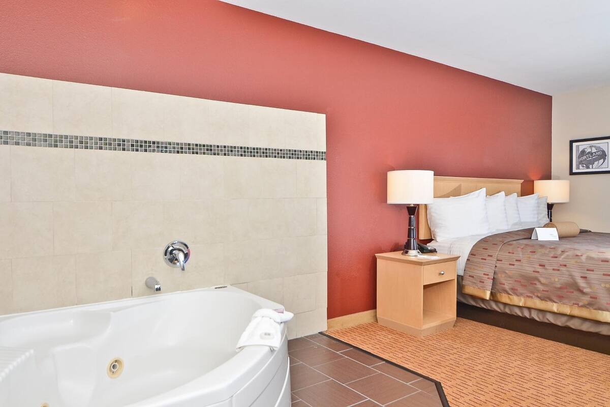 Quarto de hotel com uma banheira grande, uma cama com colcha estampada e uma mesa de cabeceira com luminárias.