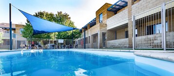 Uma piscina com toldo azul, cercada por uma vedação e um edifício com grandes janelas e fachada de tijolo.
