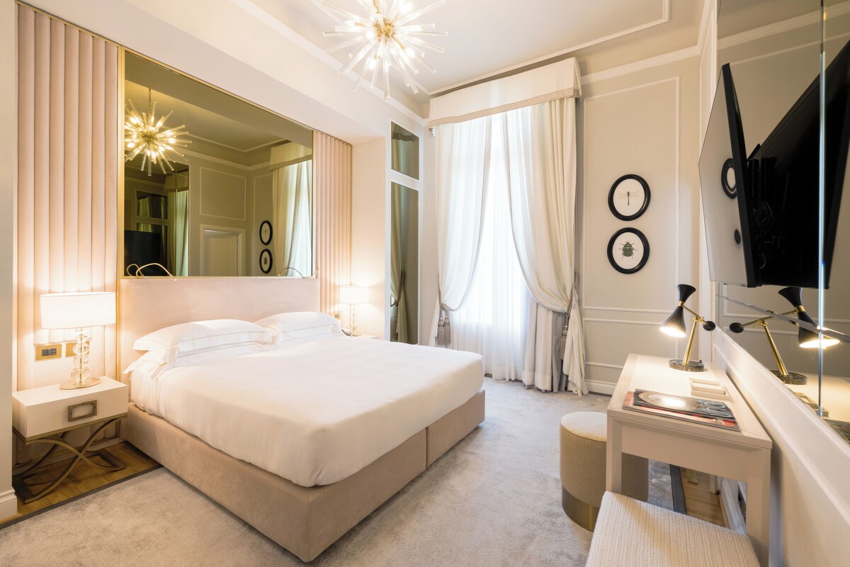 Um quarto espaçoso com uma cama grande, mesas de cabeceira, uma toucador com espelho e um lustre.