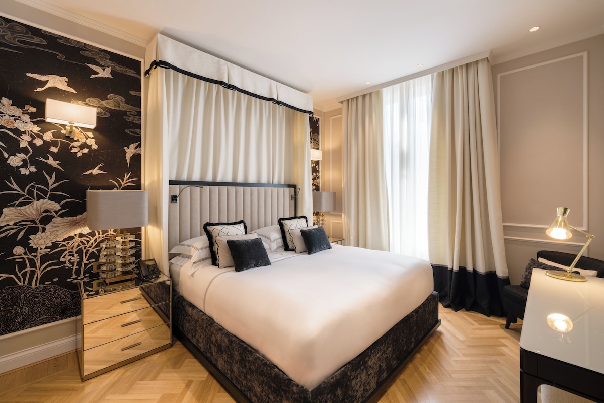 Quarto de hotel moderno com uma cama grande, mesa de cabeceira com abajur, espelho e parede decorativa com estampas florais.