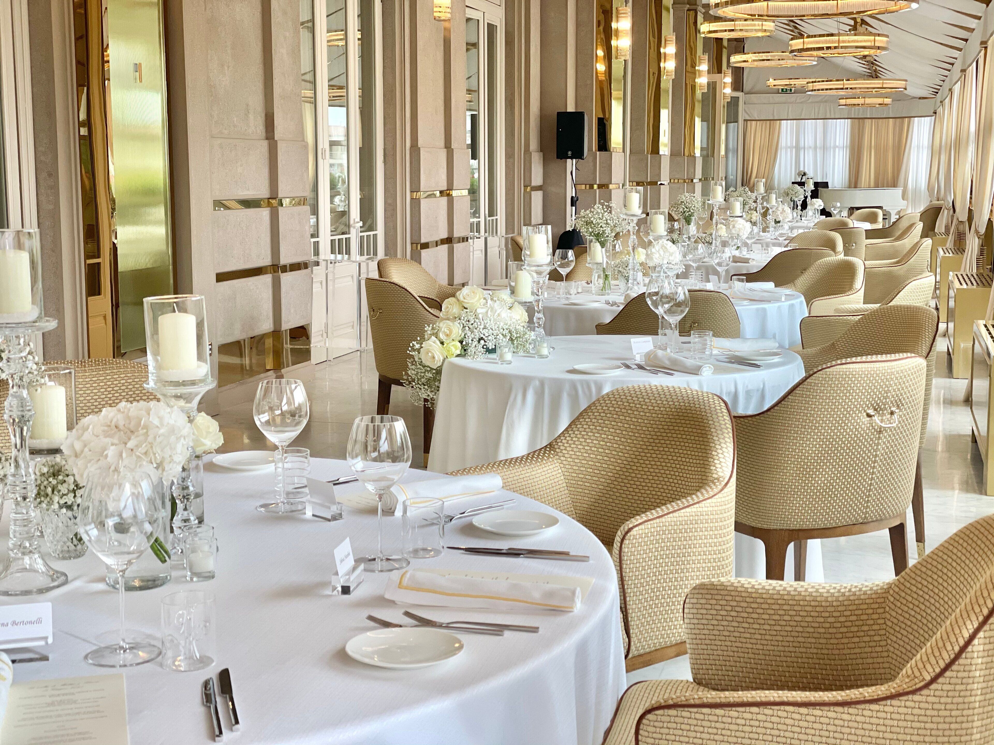 Salão de banquetes com mesas redondas dispostas para um evento formal, com toalhas brancas, copos e arranjos florais.