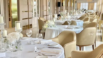 Salão de banquetes com mesas redondas dispostas para um evento formal, com toalhas brancas, copos e arranjos florais.