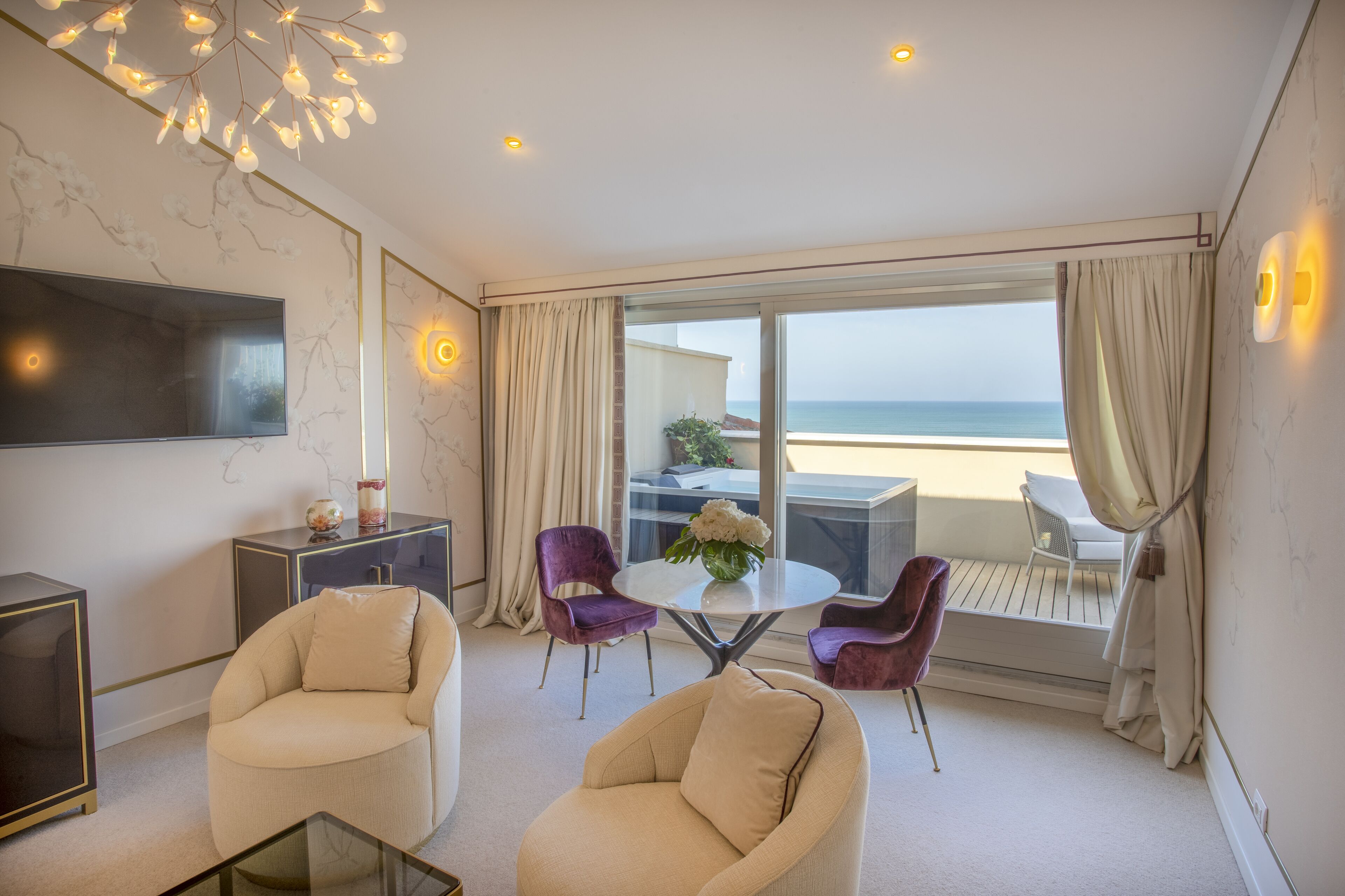 Quarto de hotel moderno com varanda, televisão de tela plana, duas poltronas, uma mesa pequena com um vaso e vista para o mar.