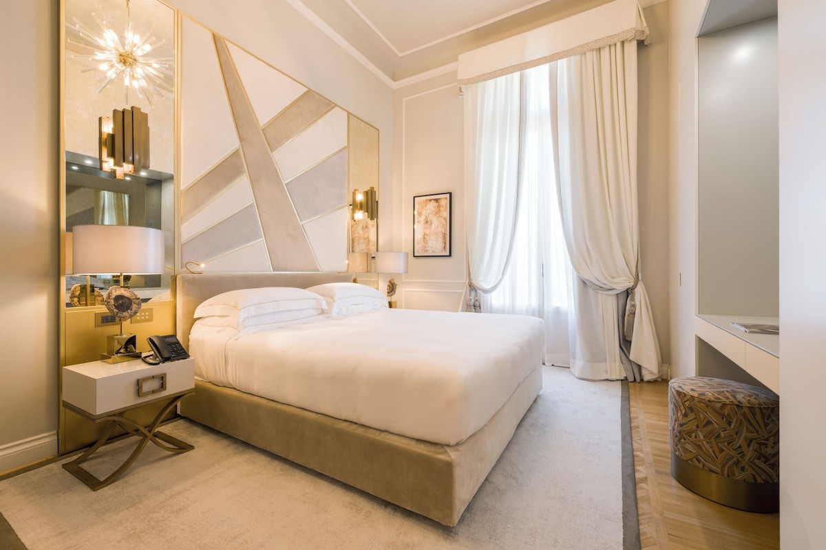 Quarto de hotel moderno com uma cama grande, mesas de cabeceira, um espelho e uma luminária decorativa.