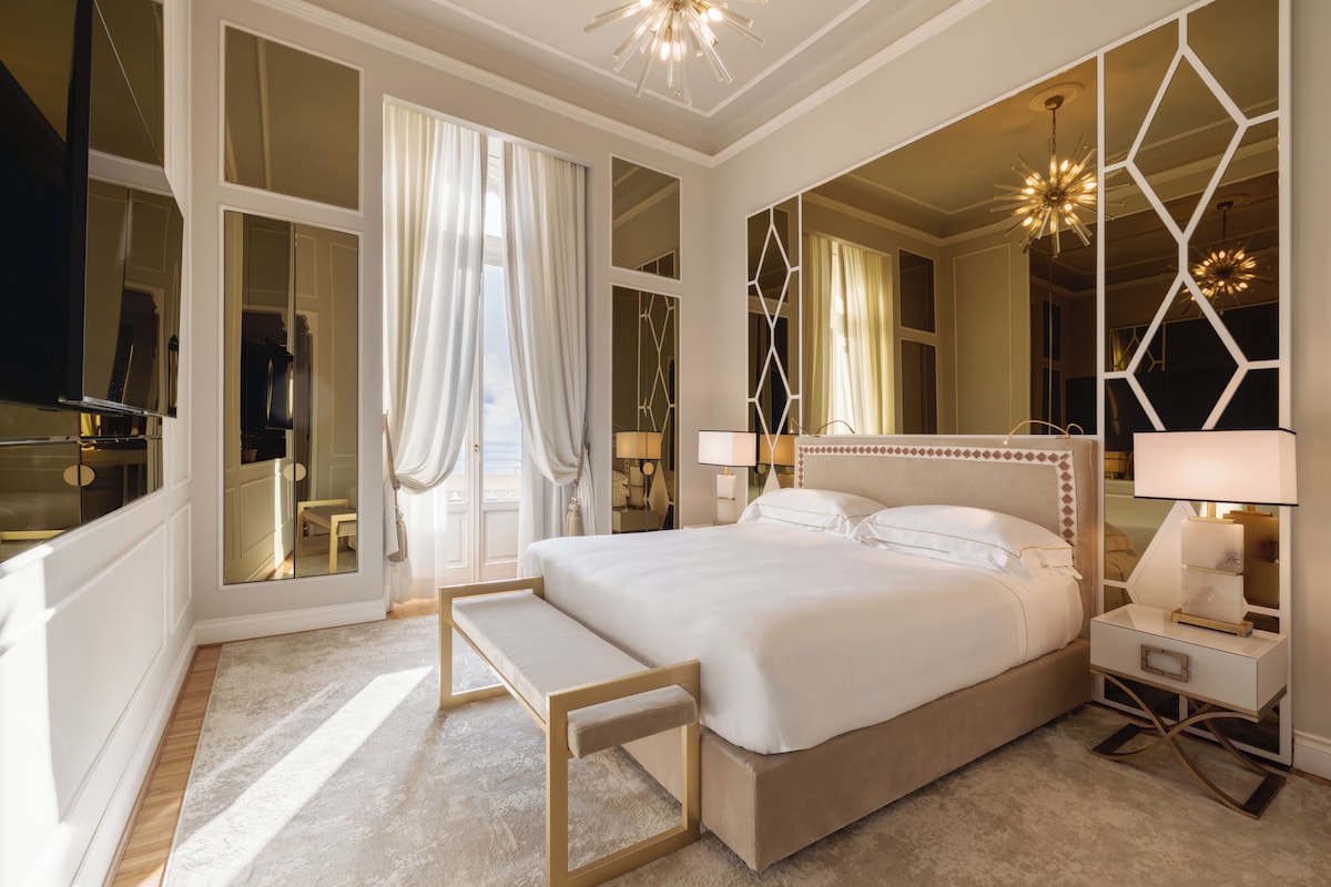 Um quarto espaçoso com uma cama grande, mesas de cabeceira, um banco e um espelho amplo.