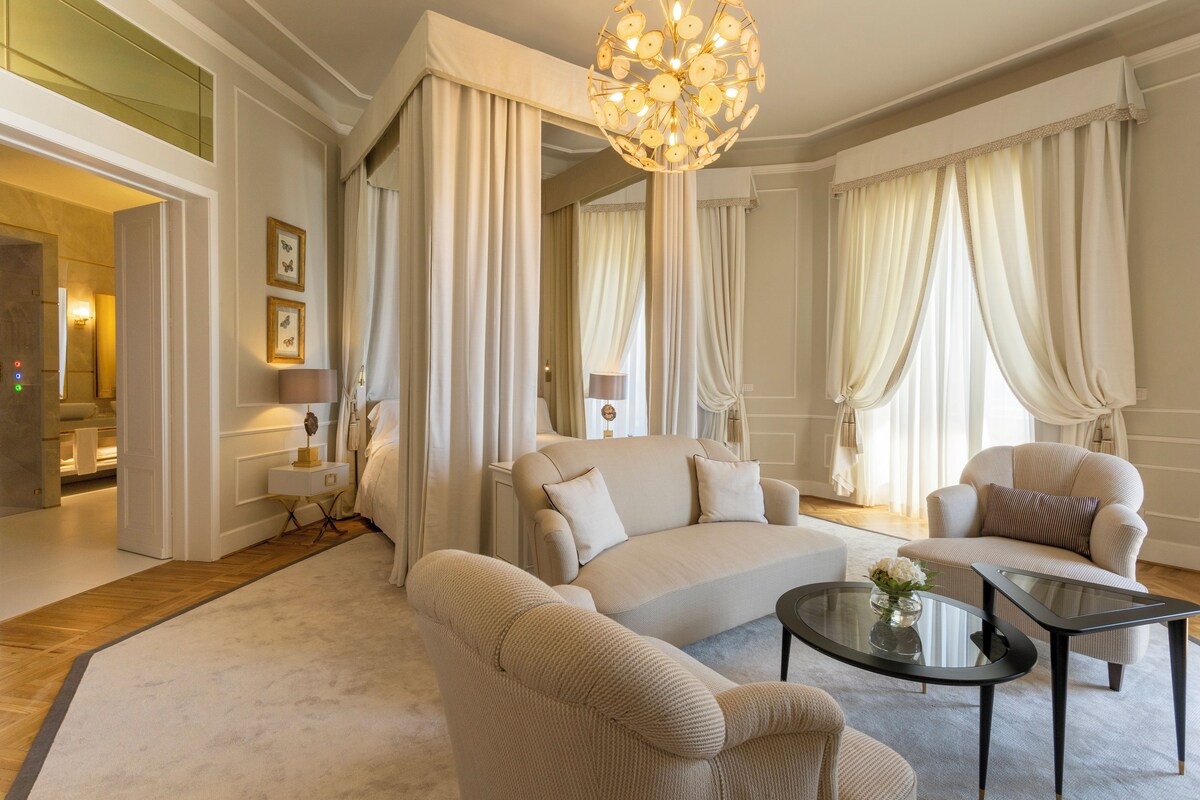 Uma sala de estar ampla com um sofá grande, uma mesa de centro e um candelabro. Há cortinas e quadros nas paredes.