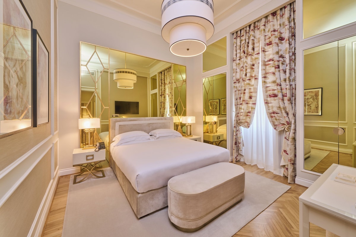 Quarto de hotel moderno com uma cama grande, uma mesa de cabeceira, um espelho e uma janela com cortinas.