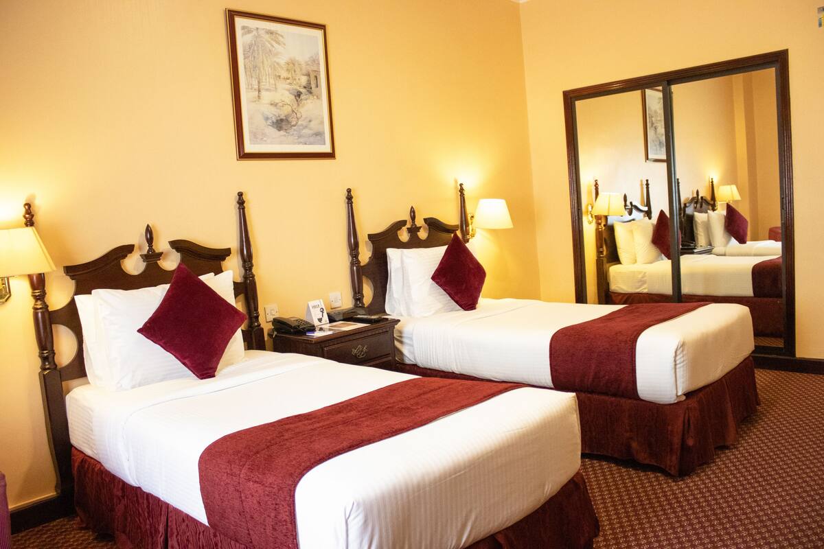 Quarto de hotel com duas camas, cada uma com lençóis brancos e um cobertor vermelho. Há uma mesinha de cabeceira com uma luminária e um telefone. Um espelho grande na parede reflete o quarto.