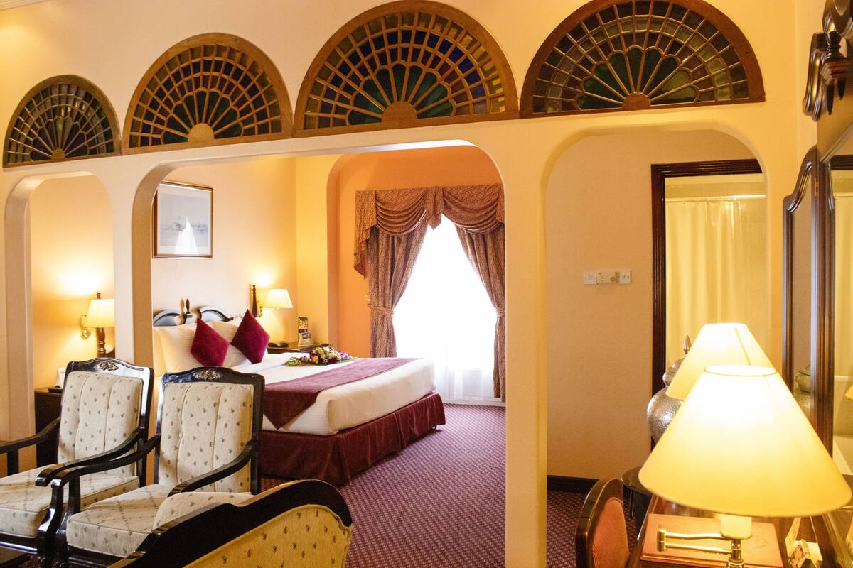 Quarto de hotel com cama, duas poltronas, mesa de cabeceira com abajur e uma janela grande com cortinas.