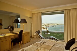 Quarto de hotel com escrivaninha, cadeira e cama. Possui uma janela grande com vista para um corpo de água e paisagem urbana.