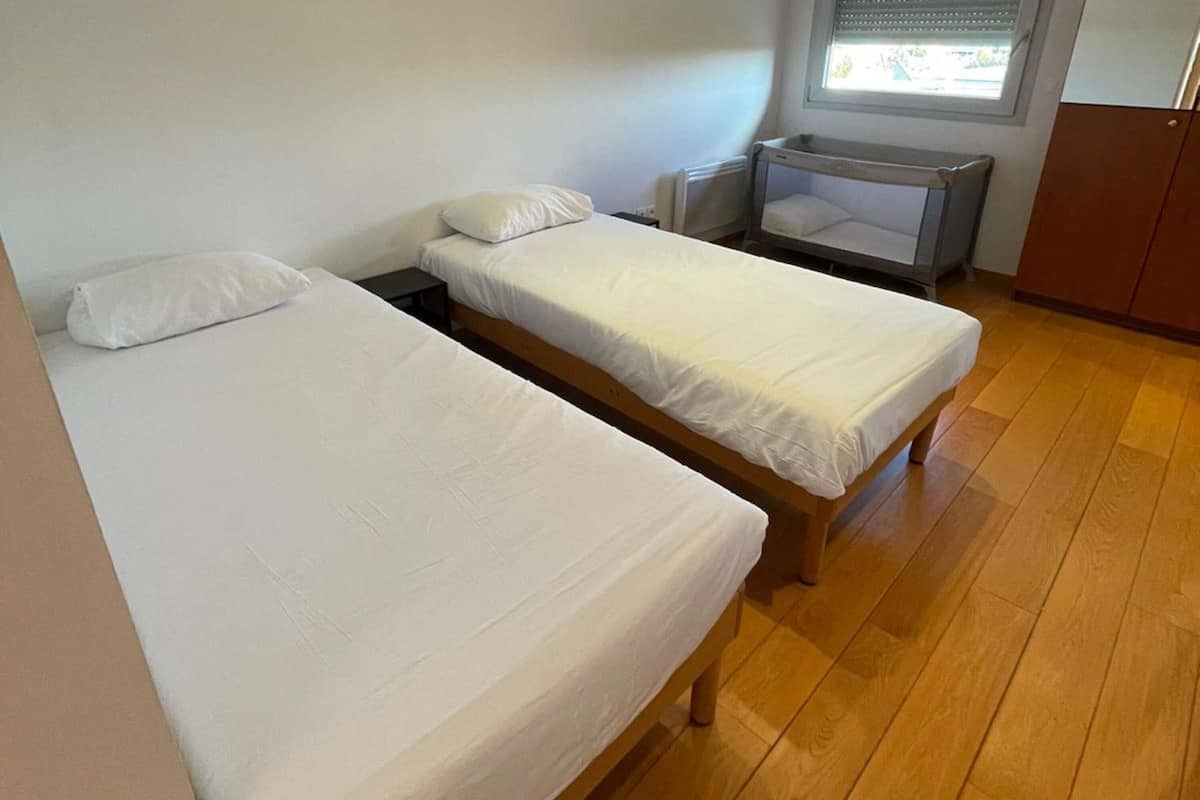 Quarto com duas camas de solteiro, uma pequena mesa de cabeceira, radiador e janela com persianas.
