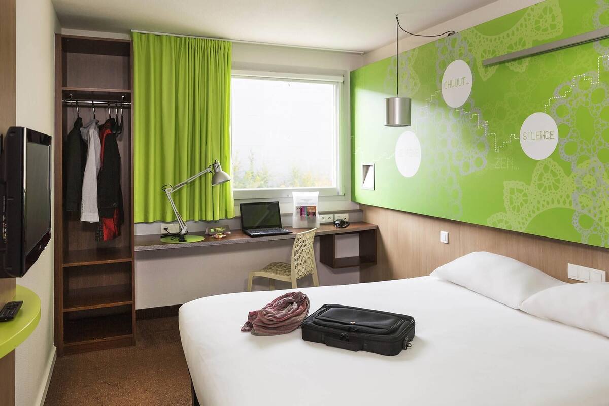 Quarto de hotel com uma cama, uma escrivaninha com um laptop, uma televisão, um guarda-roupa e uma parede verde com desenhos circulares.