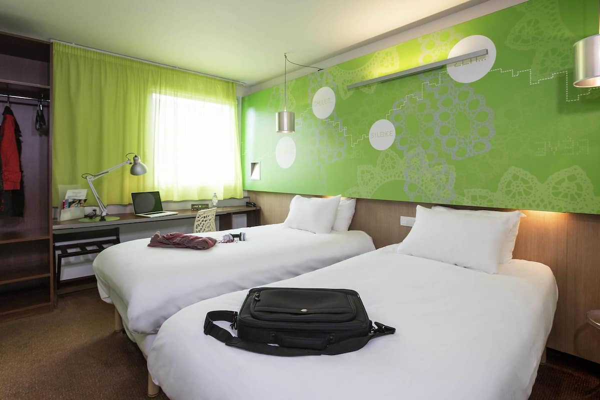 Quarto de hotel com duas camas, uma mesa com um laptop, uma luminária e uma parede verde com padrões circulares.