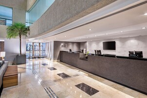 Uma área de recepção de hotel com piso de mármore, balcão de recepção, uma palmeira e uma grande janela.
