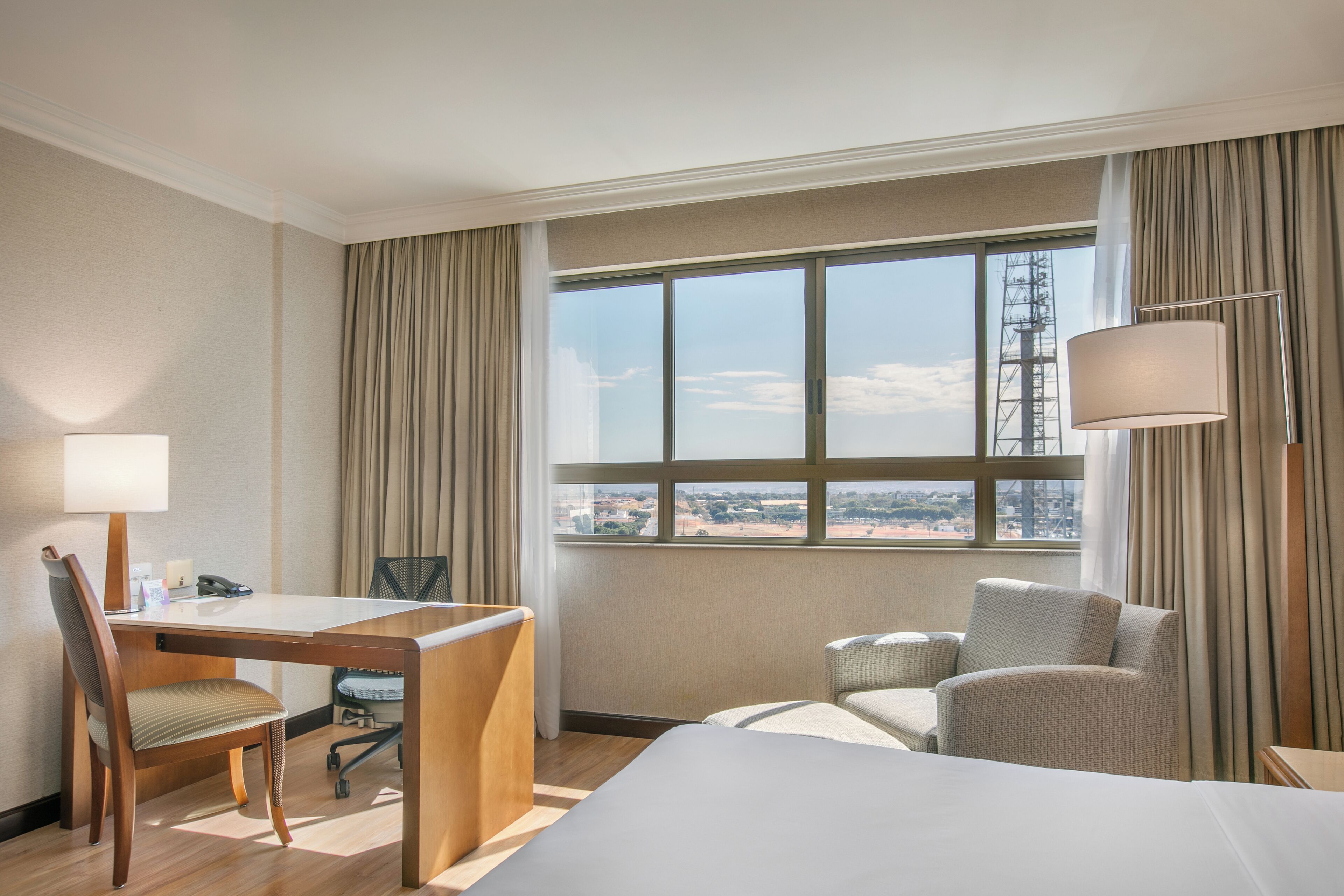 Quarto de hotel com uma cama grande, uma escrivaninha com cadeira, um sofá e vista da cidade por grandes janelas.