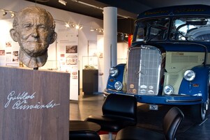 Exposição de carros antigos, com um busto em primeiro plano e painéis informativos ao fundo.