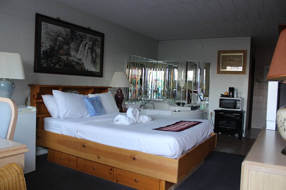 Quarto de hotel com cama de madeira, roupa de cama branca, espelho grande, televisão pequena e micro-ondas.