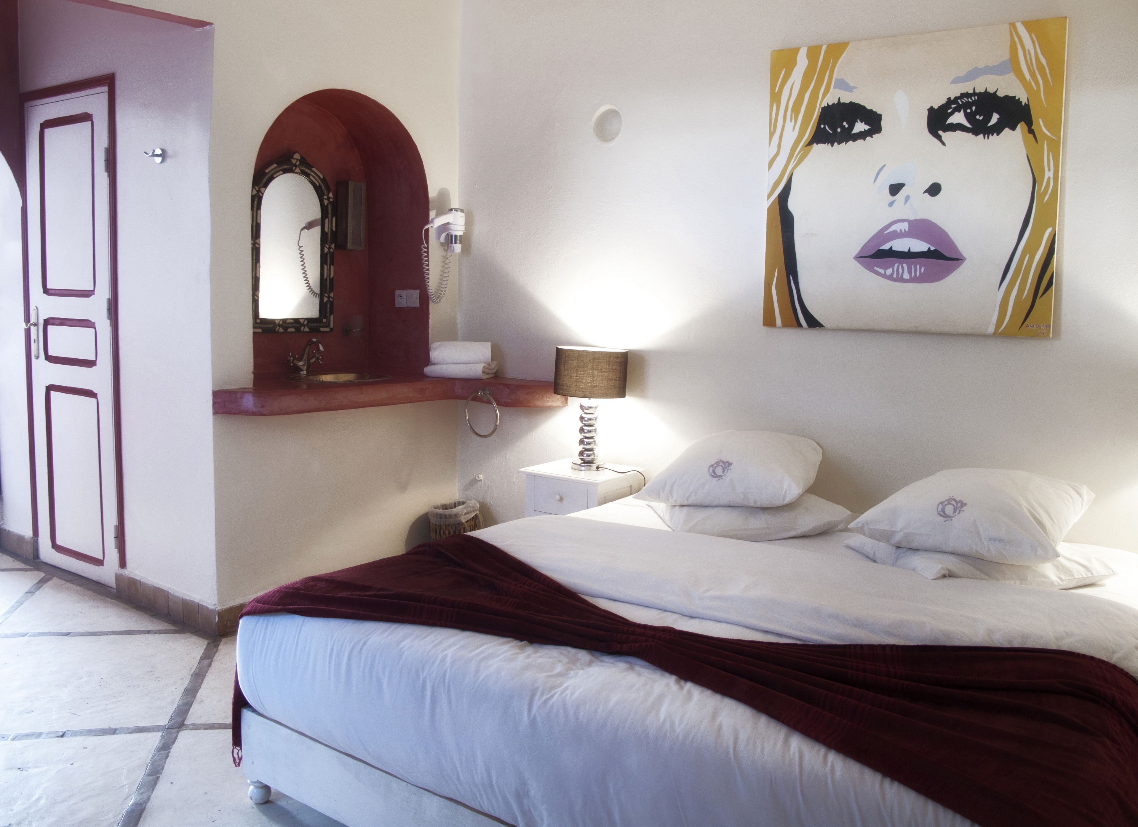 Quarto com cama, almofadas, abajur, espelho e um quadro na parede.