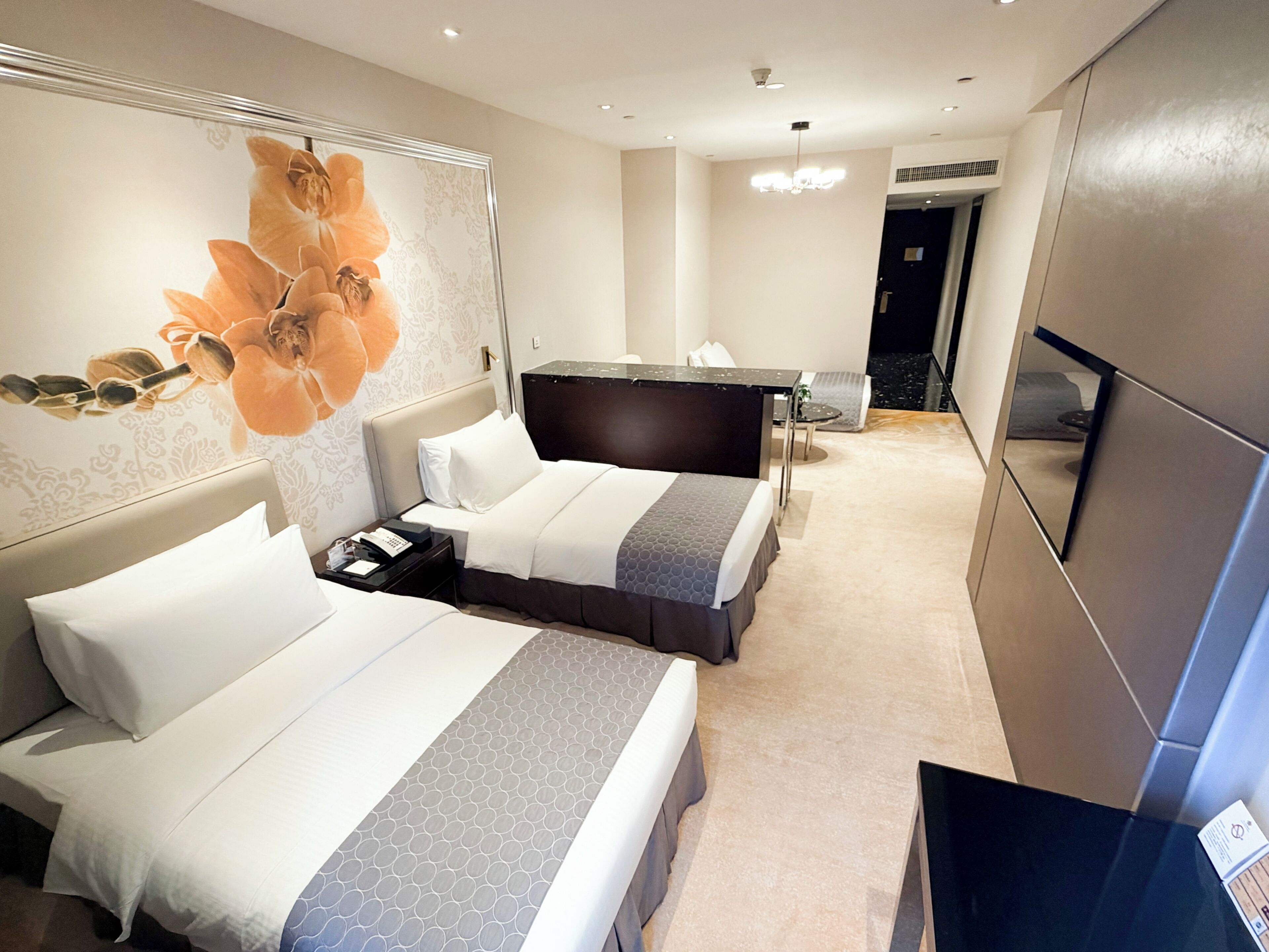 Quarto de hotel com duas camas, uma escrivaninha, uma cadeira, uma televisão e um banheiro ao fundo.