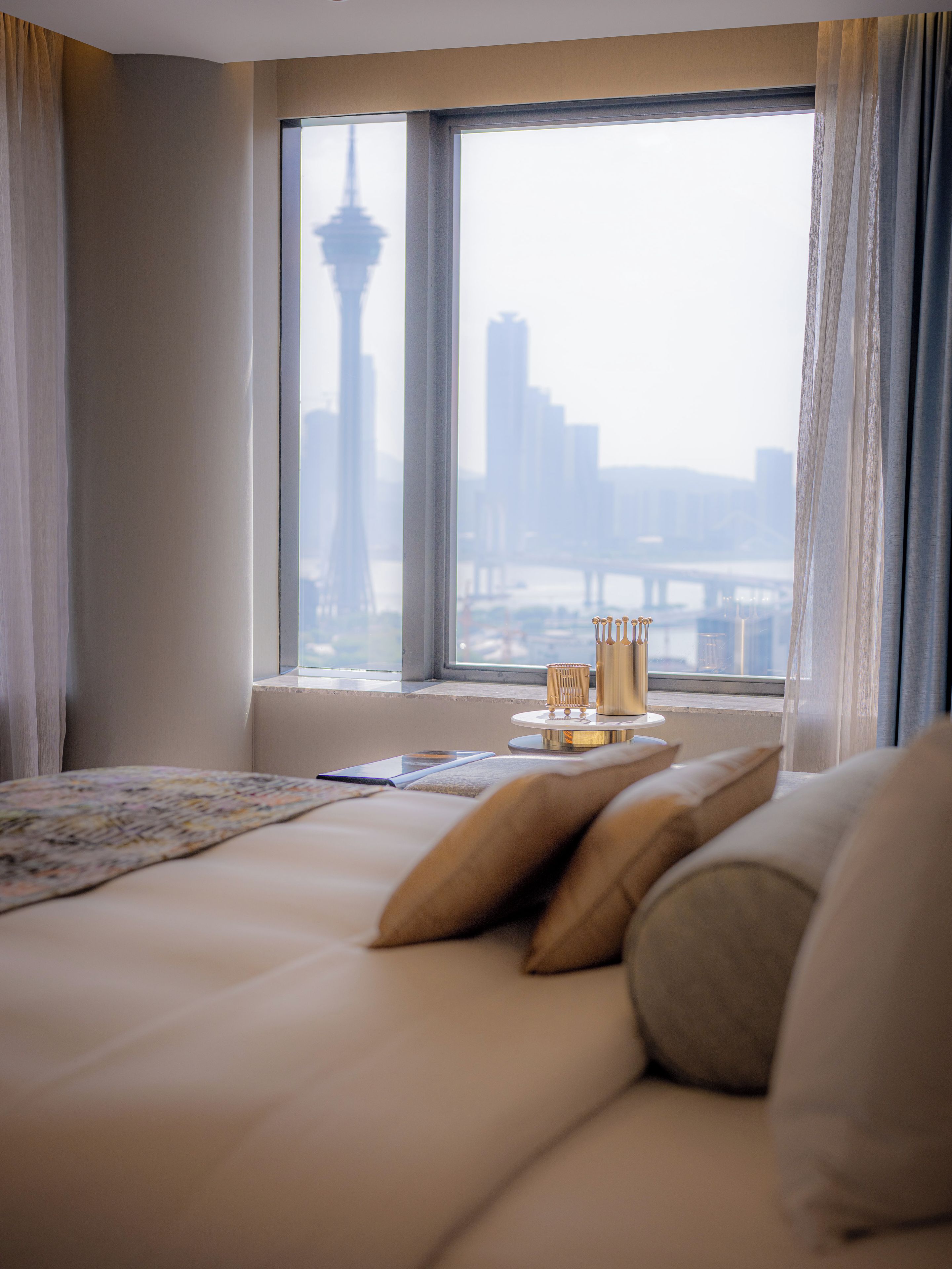 Quarto de hotel com uma grande janela oferecendo vista panorâmica da cidade, uma cama com almofadas e uma mesinha de cabeceira com abajur e um livro.