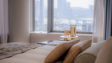 Quarto de hotel com uma grande janela oferecendo vista panorâmica da cidade, uma cama com almofadas e uma mesinha de cabeceira com abajur e um livro.