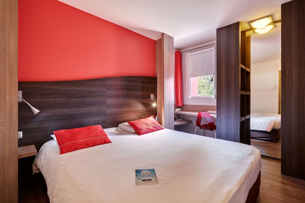Quarto de hotel com cama grande, almofadas vermelhas, mesa de cabeceira e janela com cortinas.