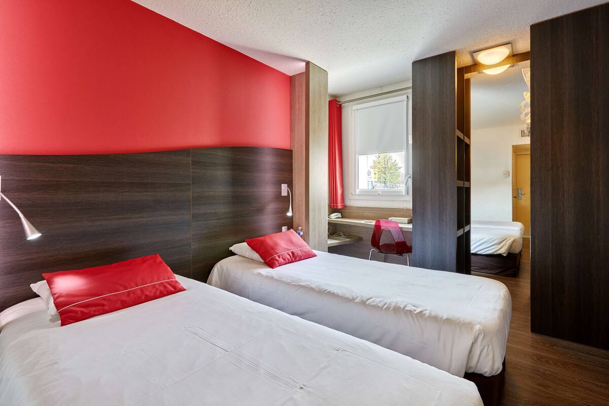 Quarto de hotel com duas camas, almofadas vermelhas, cabeceira de madeira, janela com vista e uma cadeira vermelha.