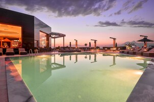 Área da piscina com espreguiçadeiras e guarda-sóis, um bar e vista para montanhas ao entardecer.