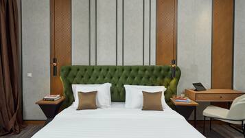 Quarto de hotel com uma cama grande, cabeceira estofada verde, duas mesas de cabeceira, uma escrivaninha e uma cadeira.