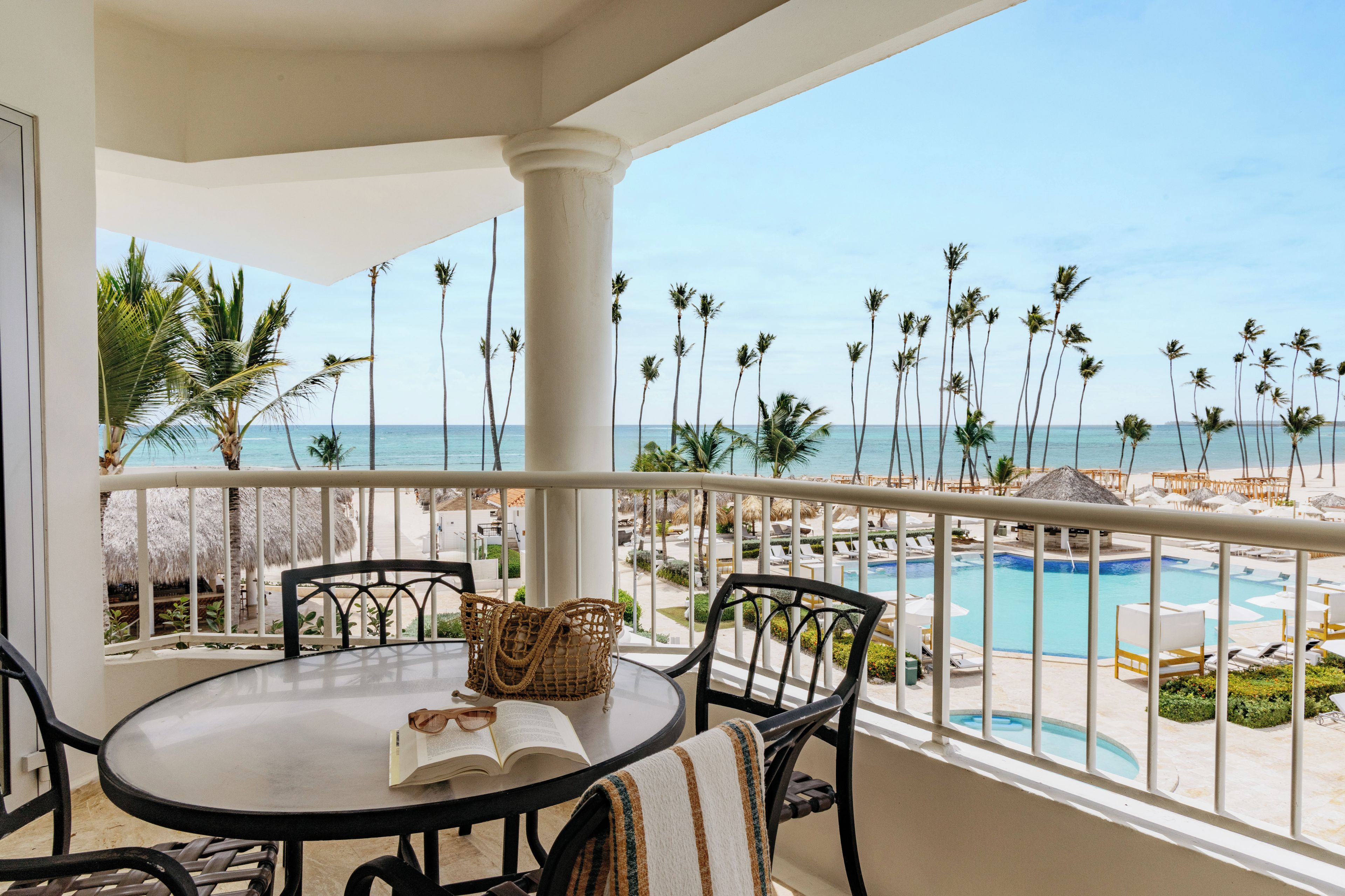 Varanda com vista para a praia e piscina, mobiliada com mesa, cadeiras e um cesto de vime.