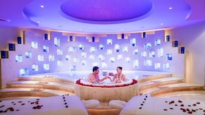 Um casal em um jacuzzi circular rodeado por pétalas de rosa, com um painel iluminado ao fundo.
