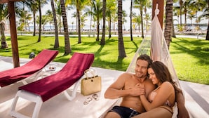 Um casal relaxando em uma rede, em uma área de cadeiras de praia, com vista para palmeiras e o oceano.