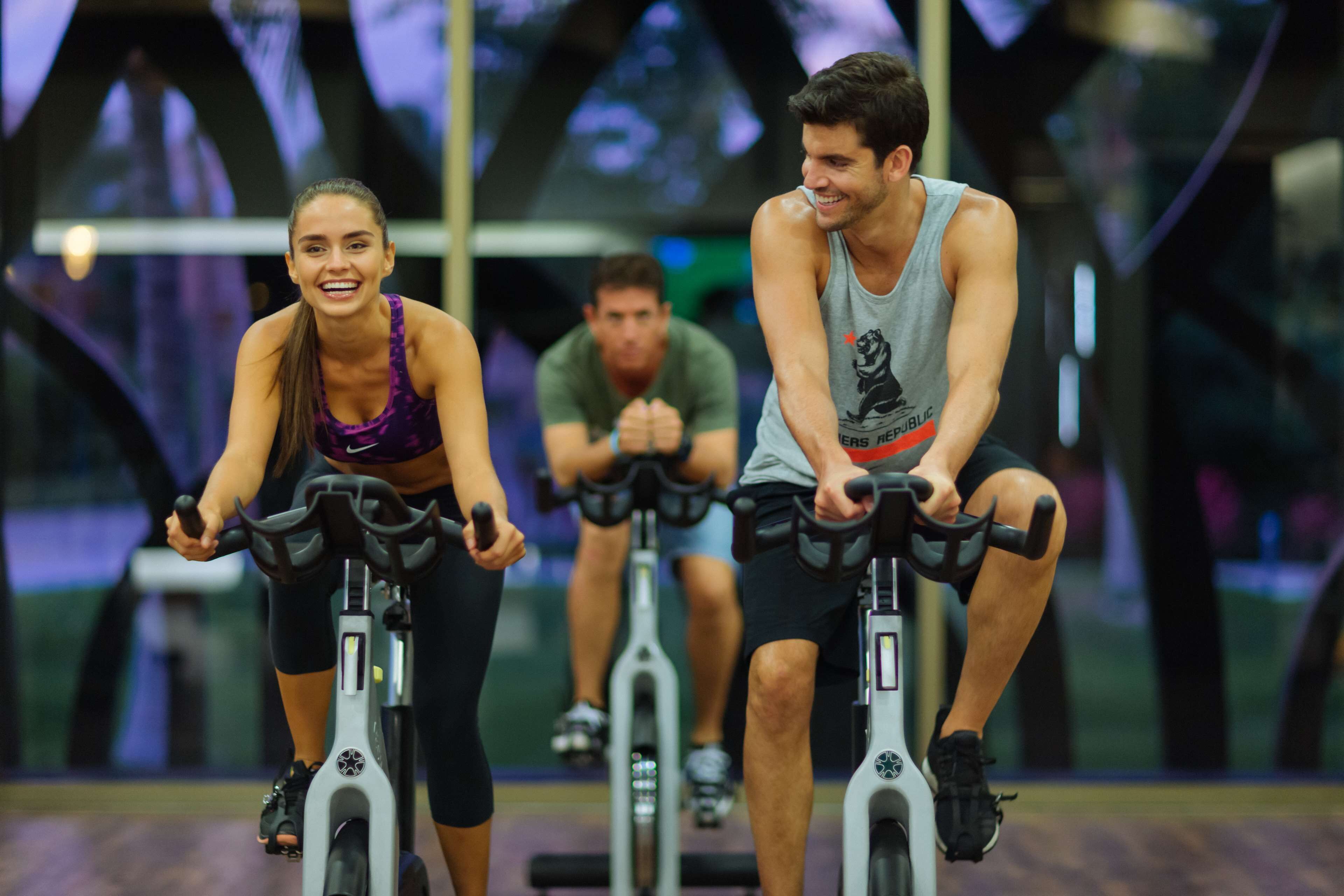 Três pessoas pedalando bicicletas ergométricas numa academia com paredes espelhadas e design moderno.